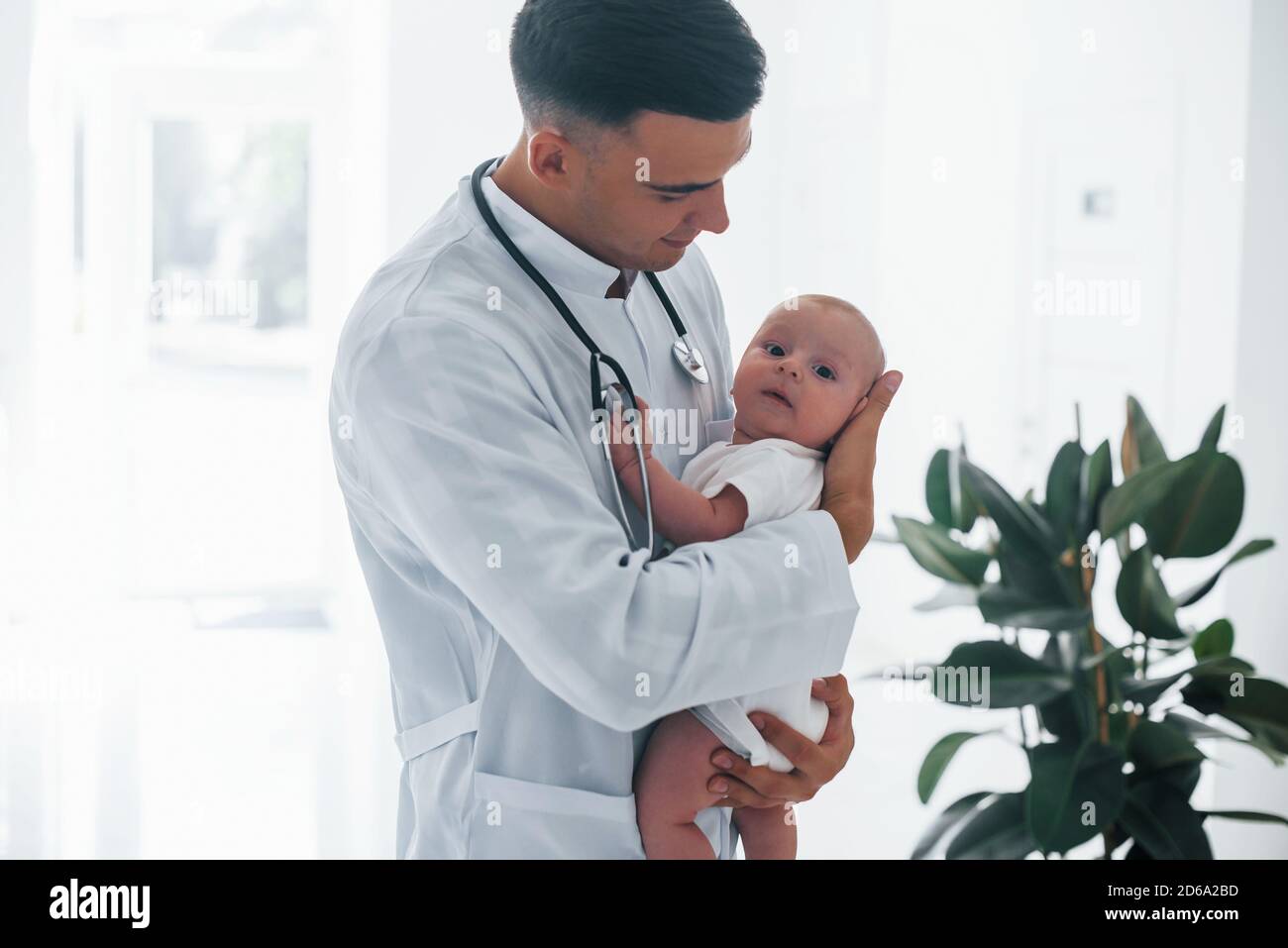 Stands against window. Young pediatrician is with little baby in the ...
