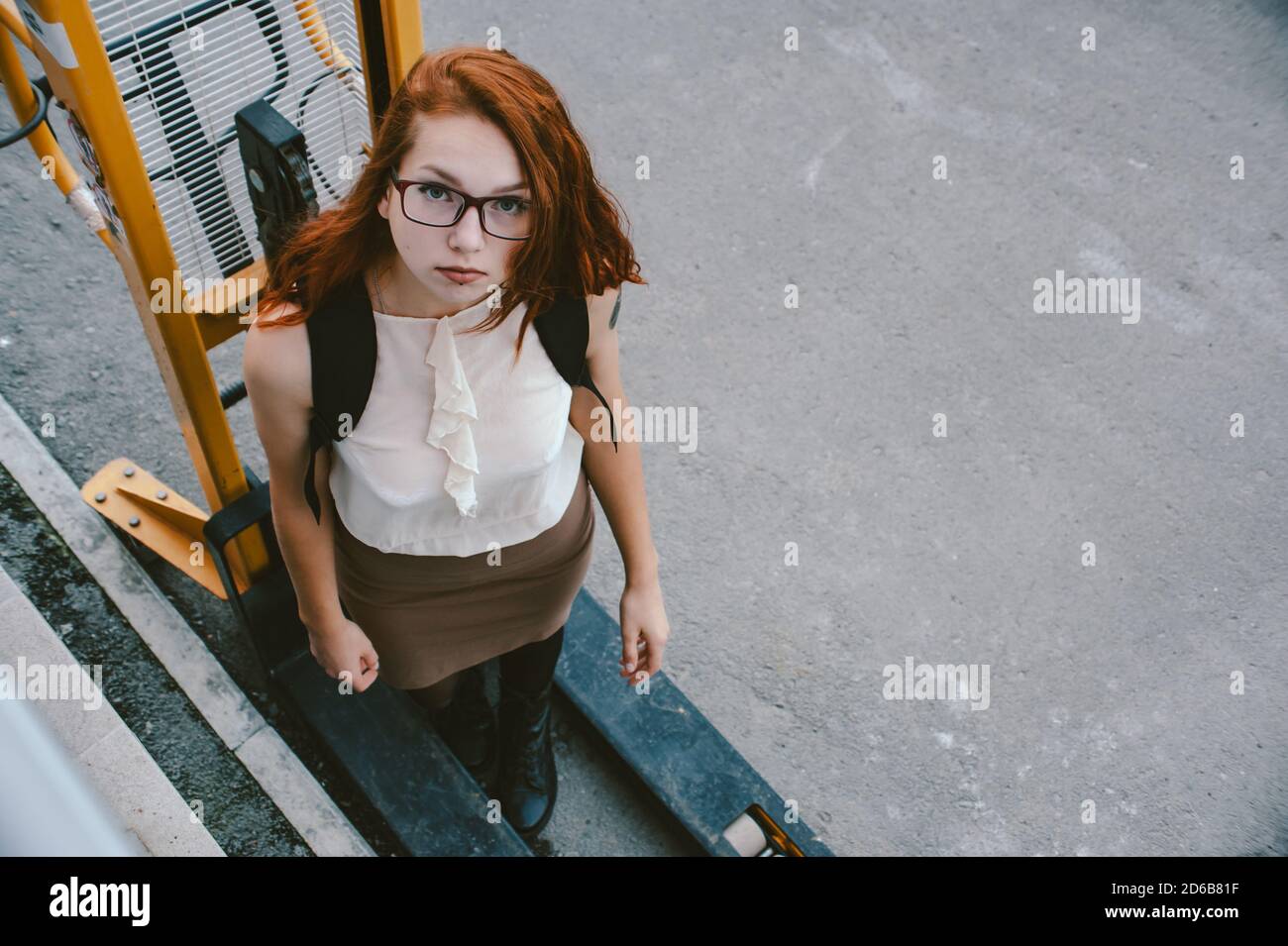 A girl in light clothes and red hair stands on a hydraulic stackers ...