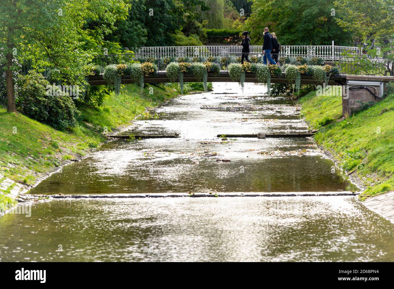 People strolling over a pedestrian bridge over a small canal in a park ...