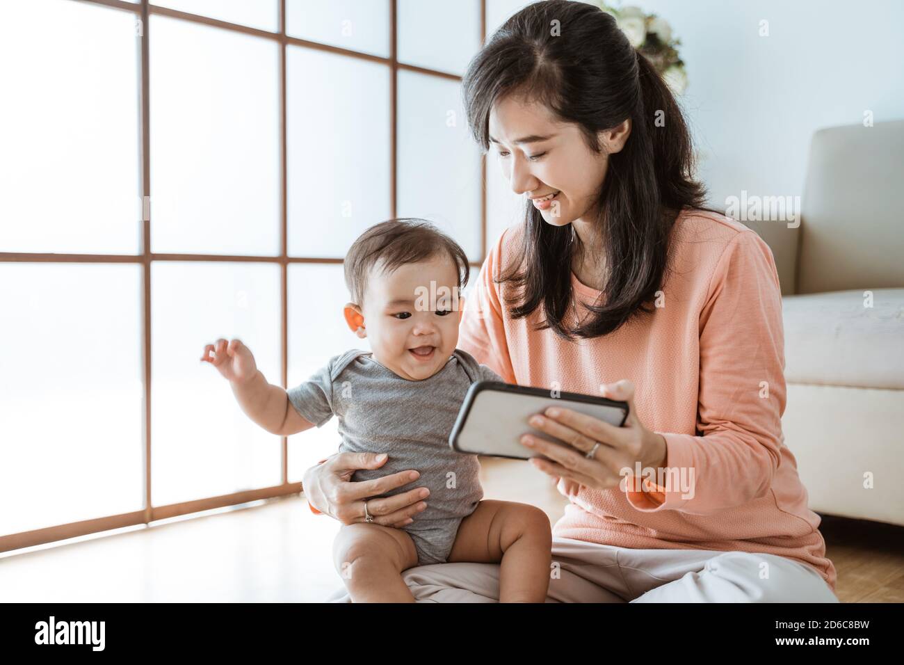 mother using mobile phone with her baby while sitting at home living ...