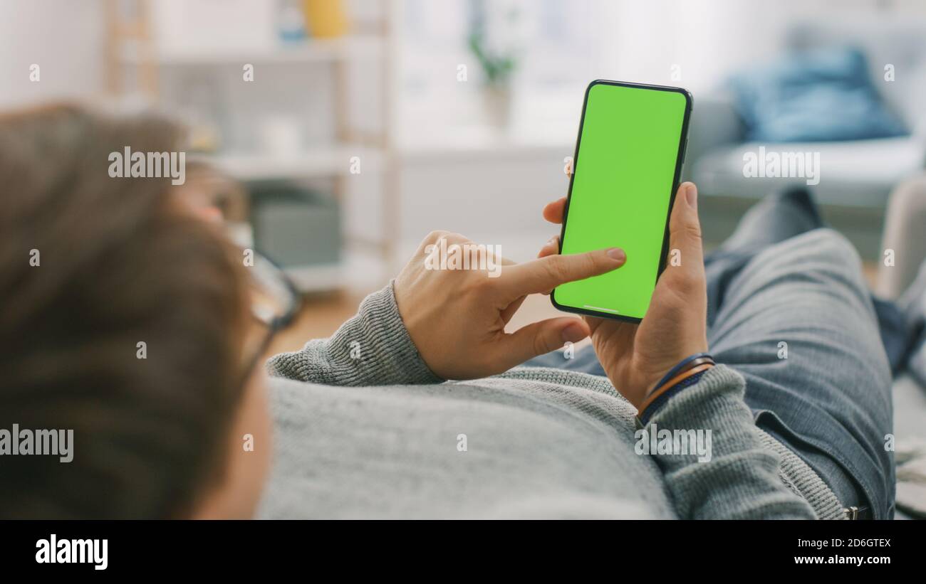 Man at Home Lying on a Couch using Smartphone with Green Mock-up Screen, Doing Swiping ...