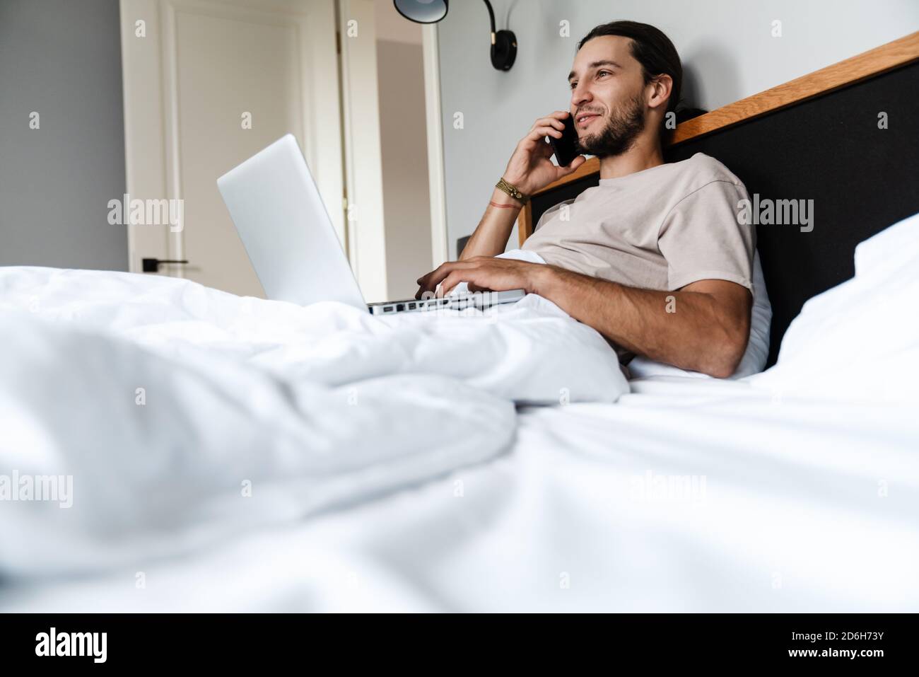 Smiling young man using mobile phone and laptop computer while laying ...