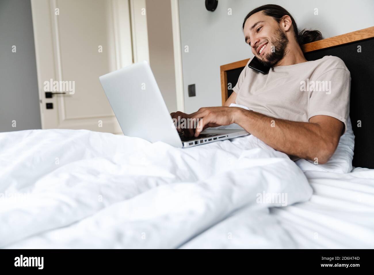 Smiling young man using mobile phone and laptop computer while laying ...
