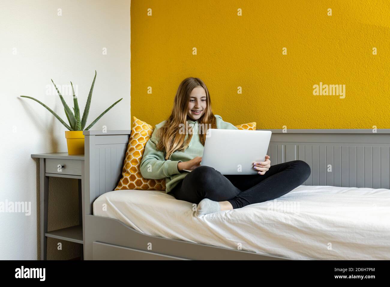 Young blonde girl using laptop computer on her bed in fornt of a yellow ...