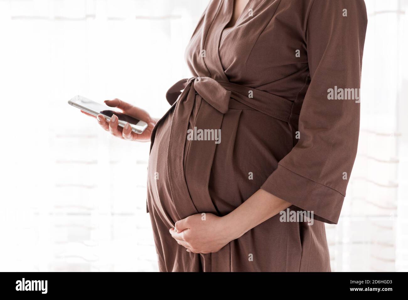 Pretty young pregnant woman standing by the window and texting message ...
