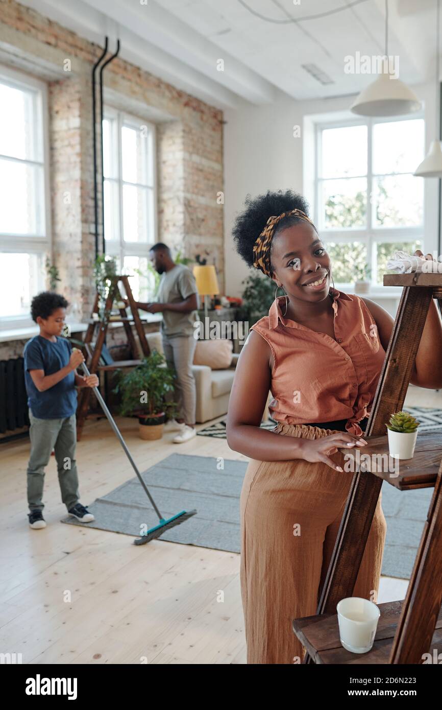 Young African female cleaning wooden shelves with duster in front of ...