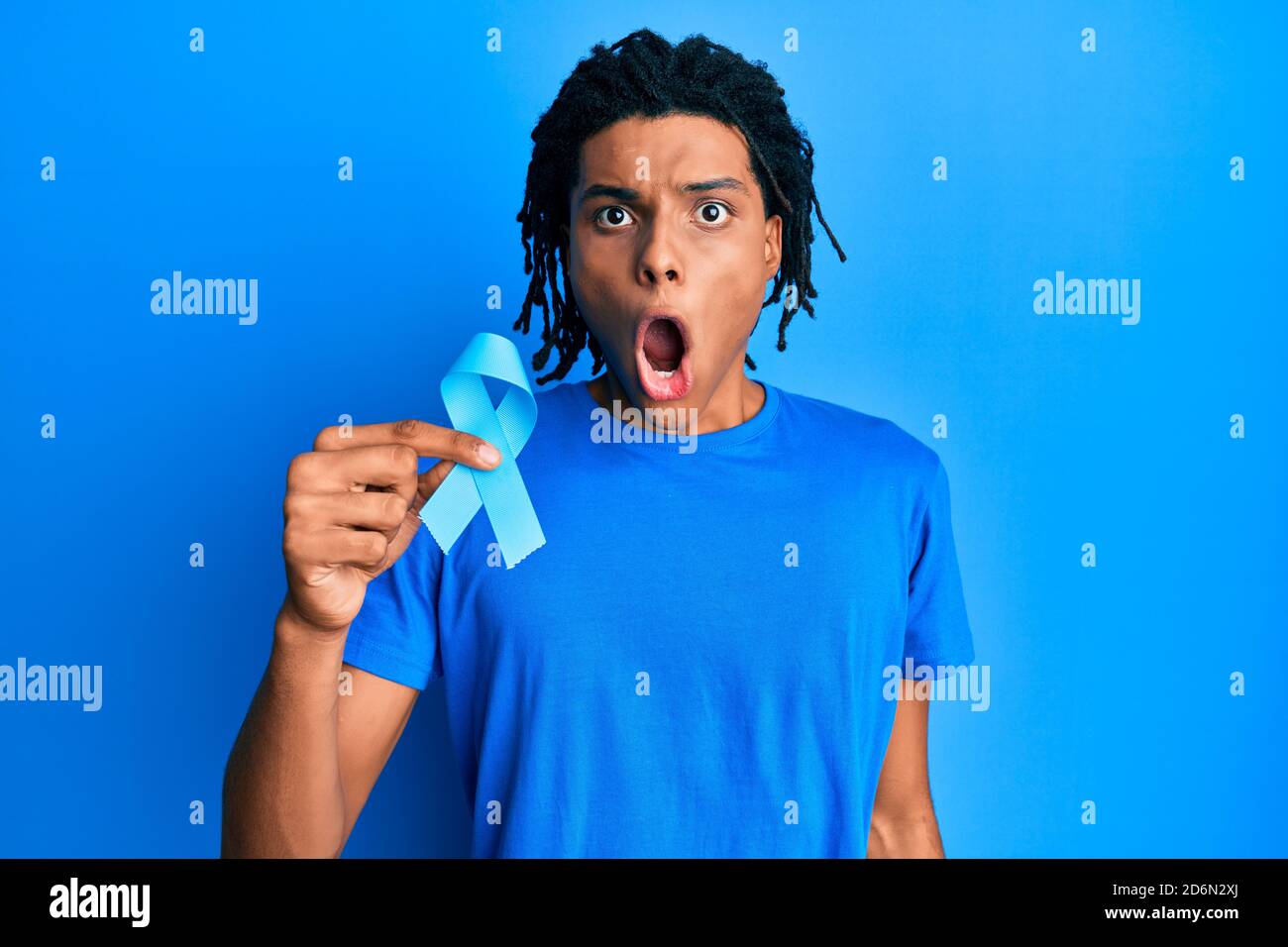 Young african american man holding blue ribbon scared and amazed with ...