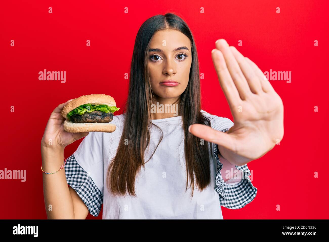 Young hispanic woman eating hamburger with open hand doing stop sign ...