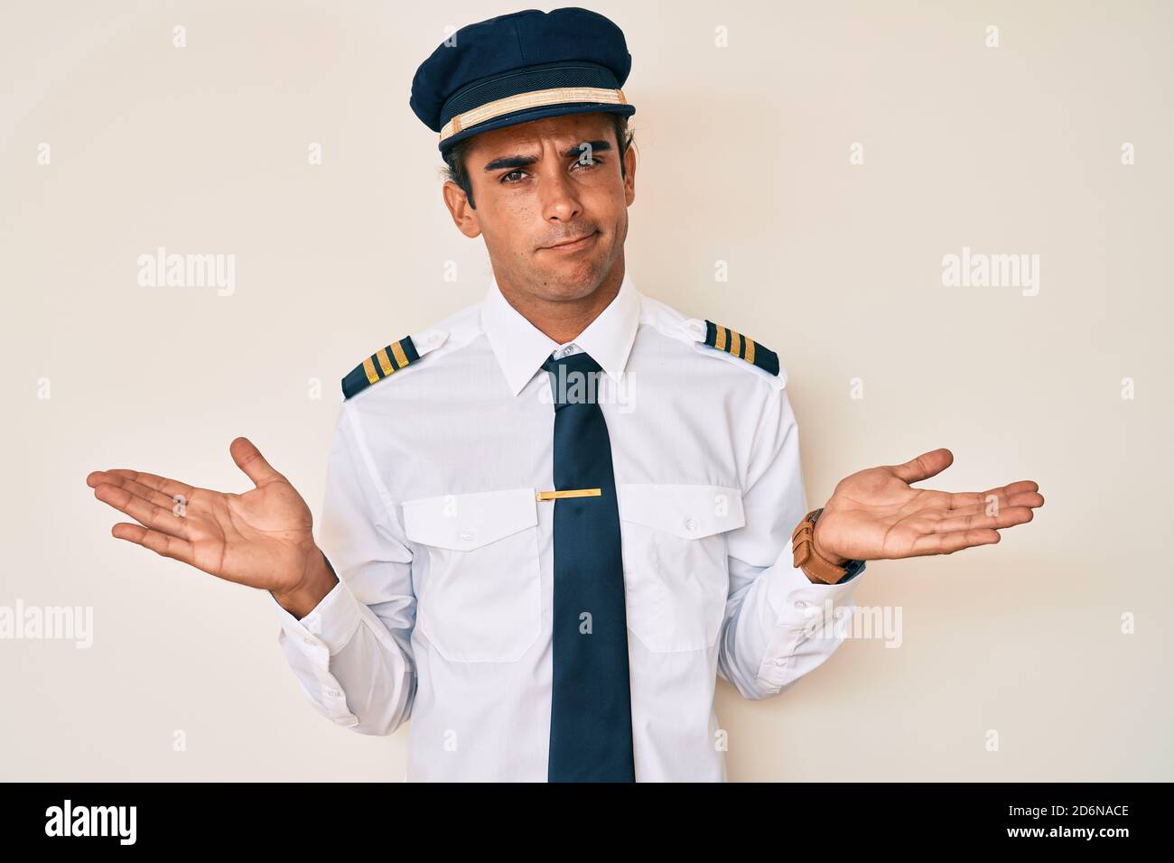 Young hispanic man wearing airplane pilot uniform clueless and confused ...