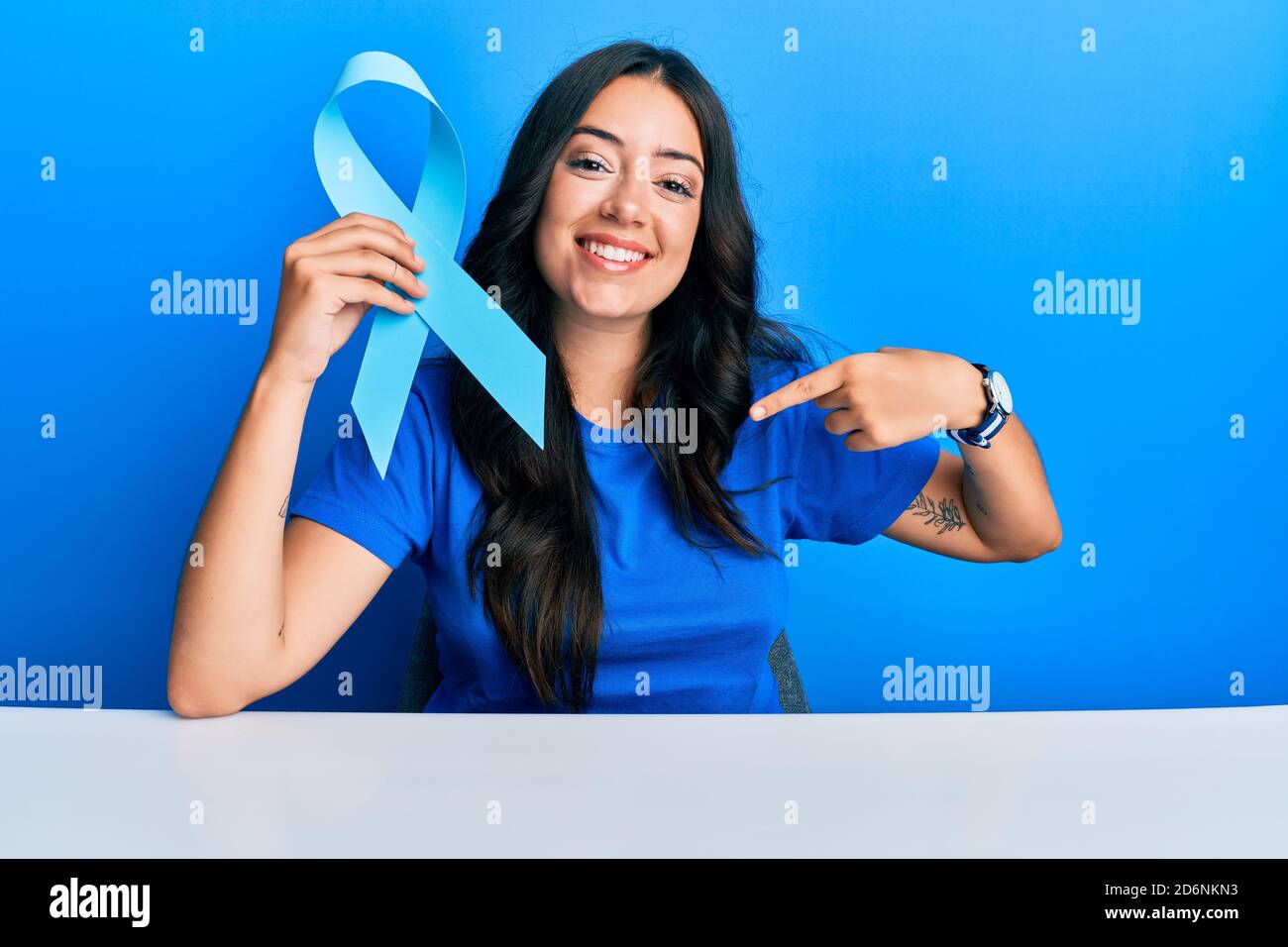 Beautiful brunette young woman holding blue ribbon pointing finger to ...