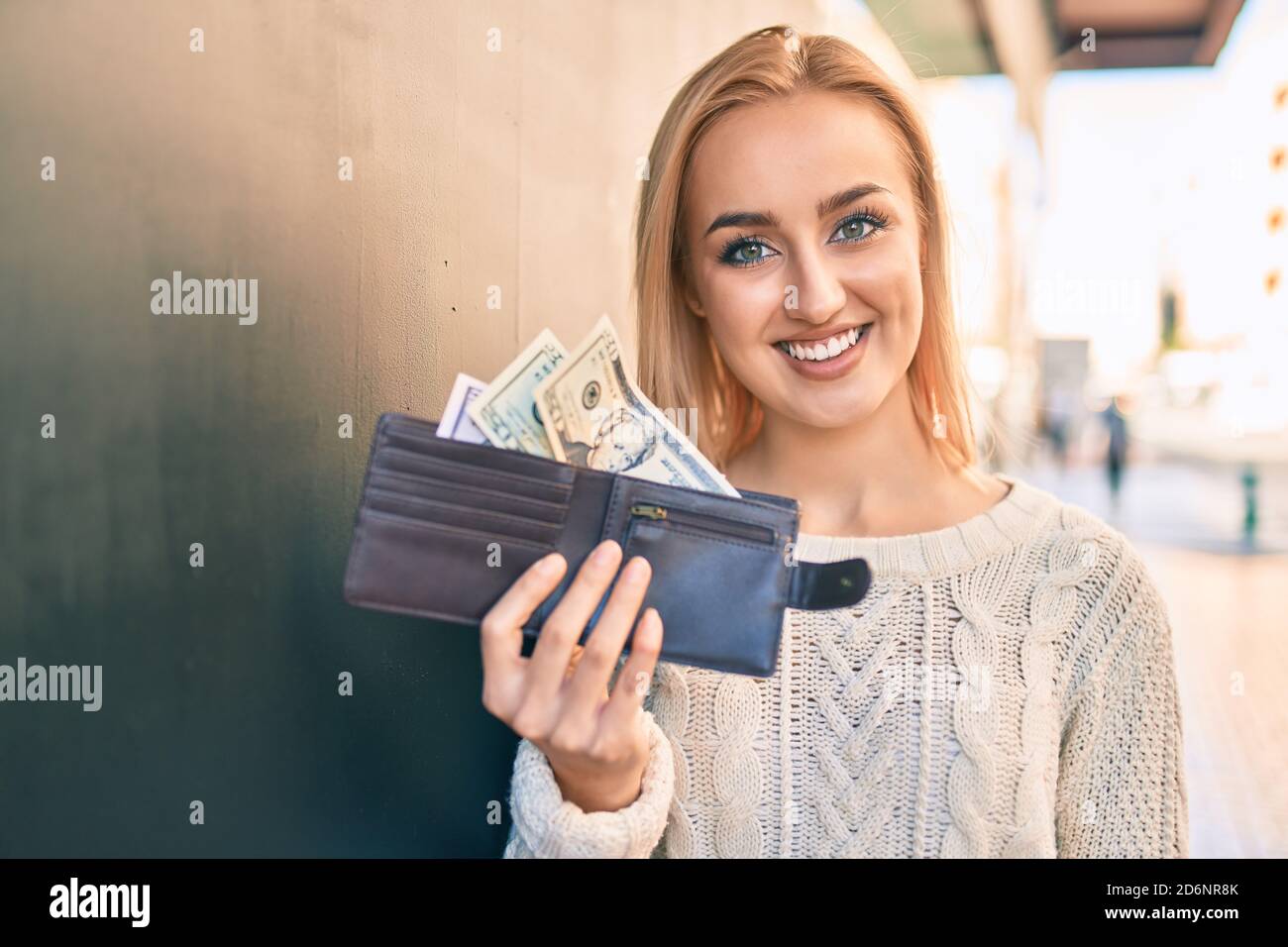 Young blonde girl smiling happy holding wallet with dollars standing at ...