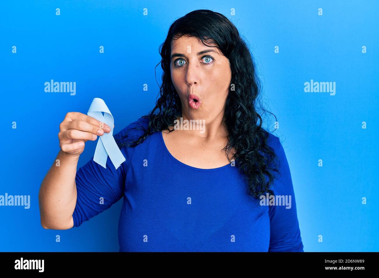 Plus size brunette woman holding blue ribbon scared and amazed with ...