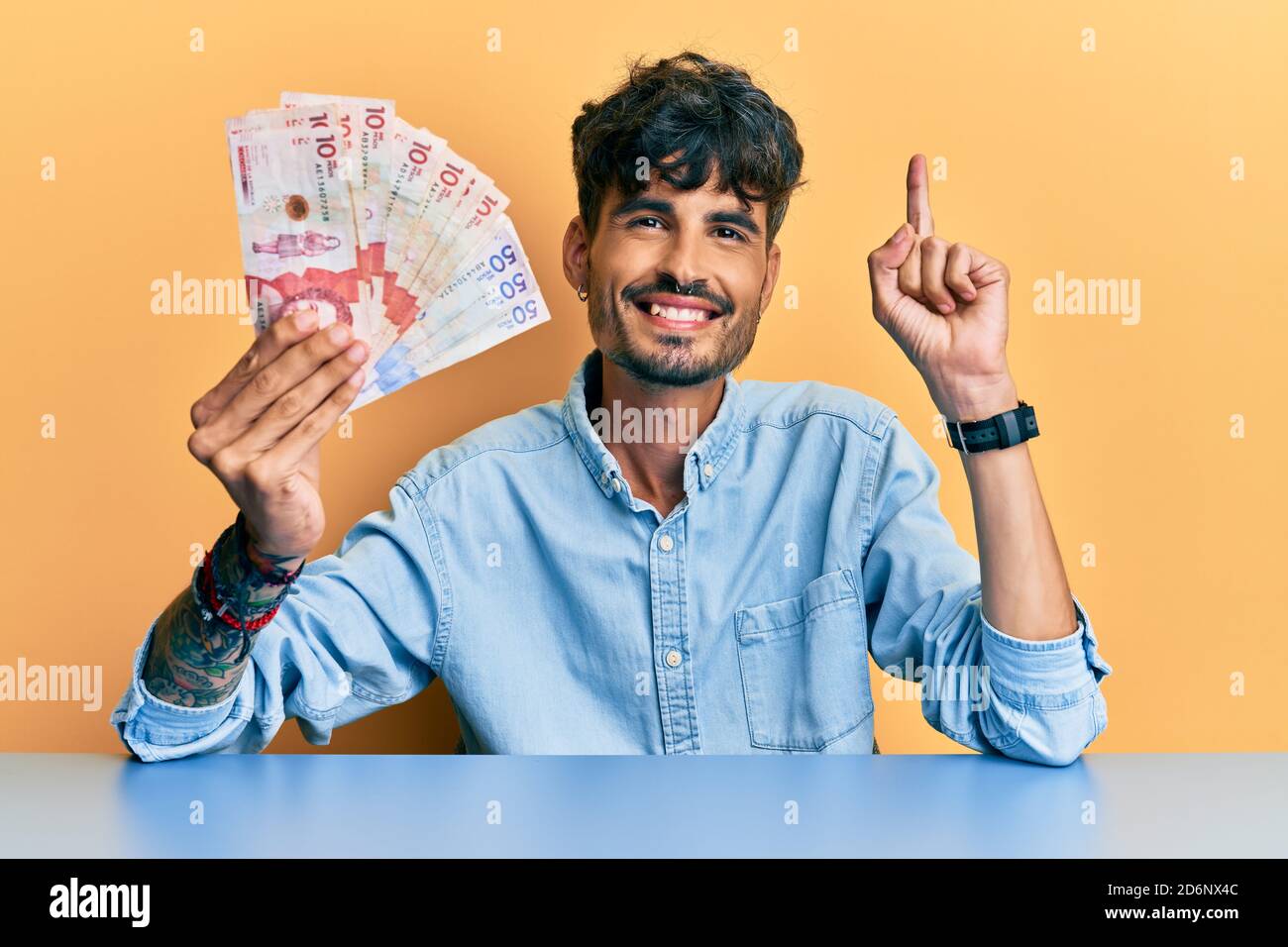 Young hispanic man holding colombian pesos sitting on the table smiling ...