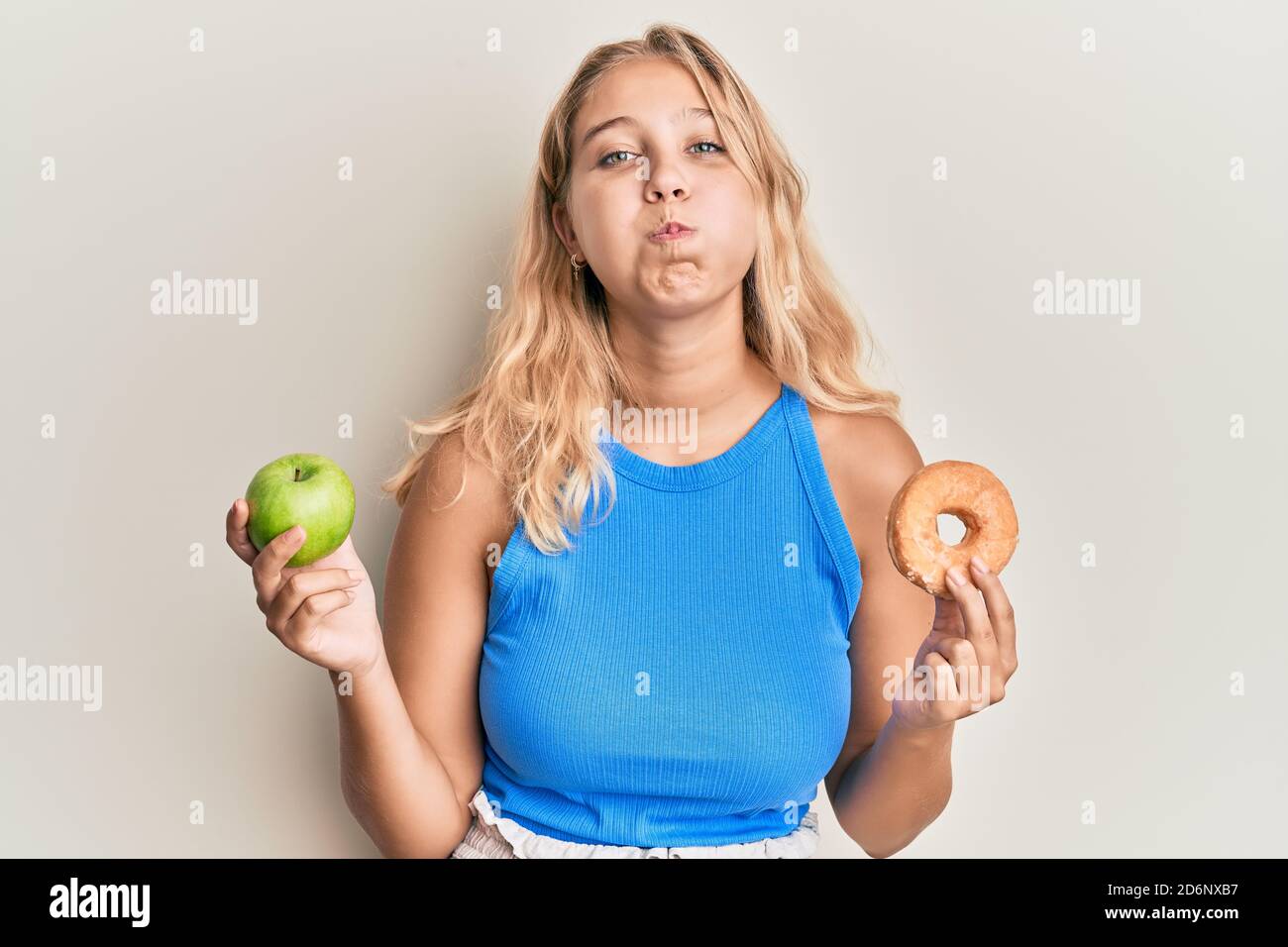 Young blonde girl holding green apple and donut puffing cheeks with ...