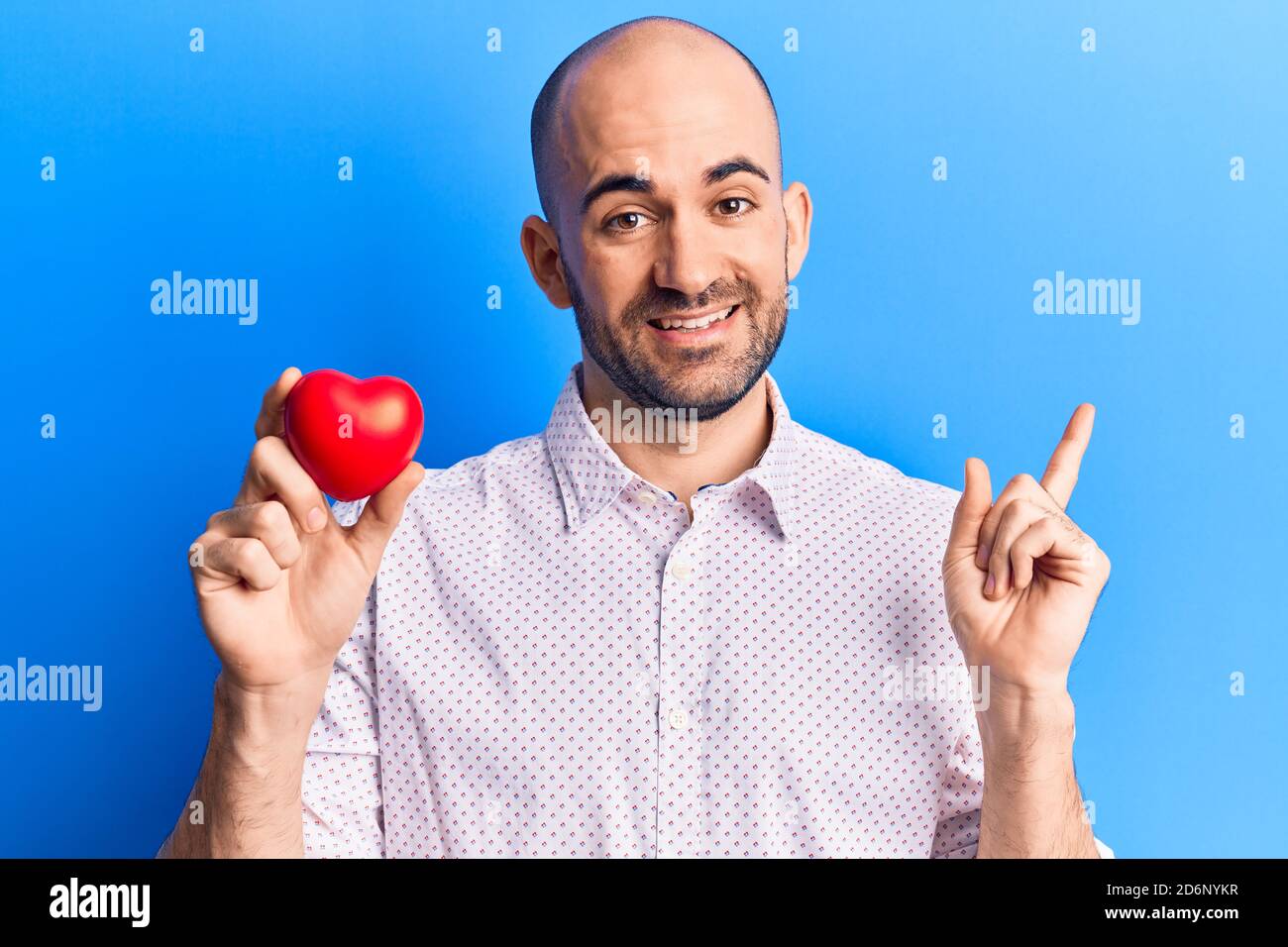 Young handsome bald man holding heart smiling happy pointing with hand ...