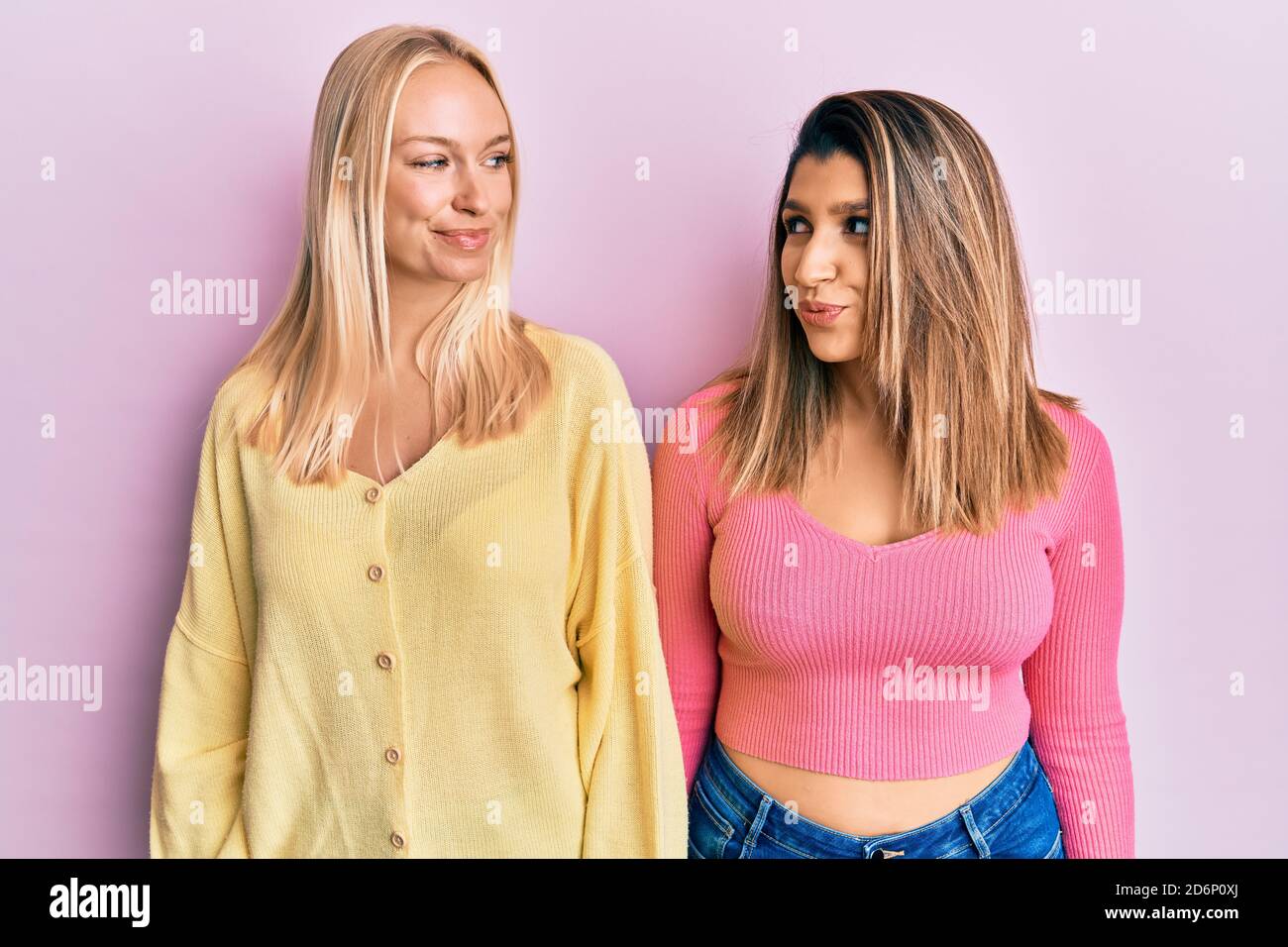 Two friends standing together over pink background smiling looking to ...