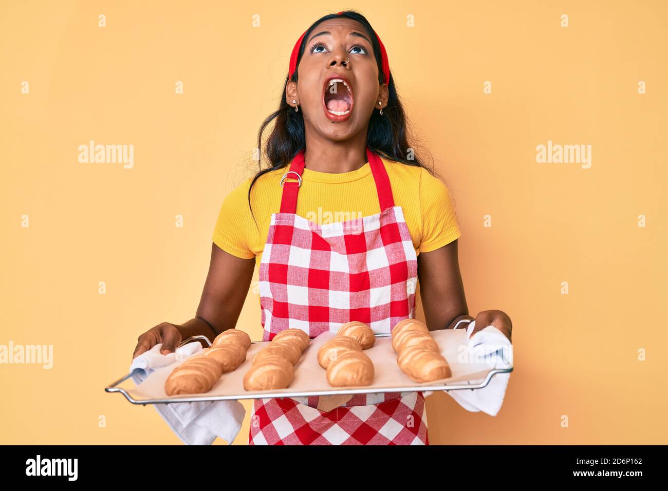 Young indian girl wearing baker uniform holding homemade bread angry ...