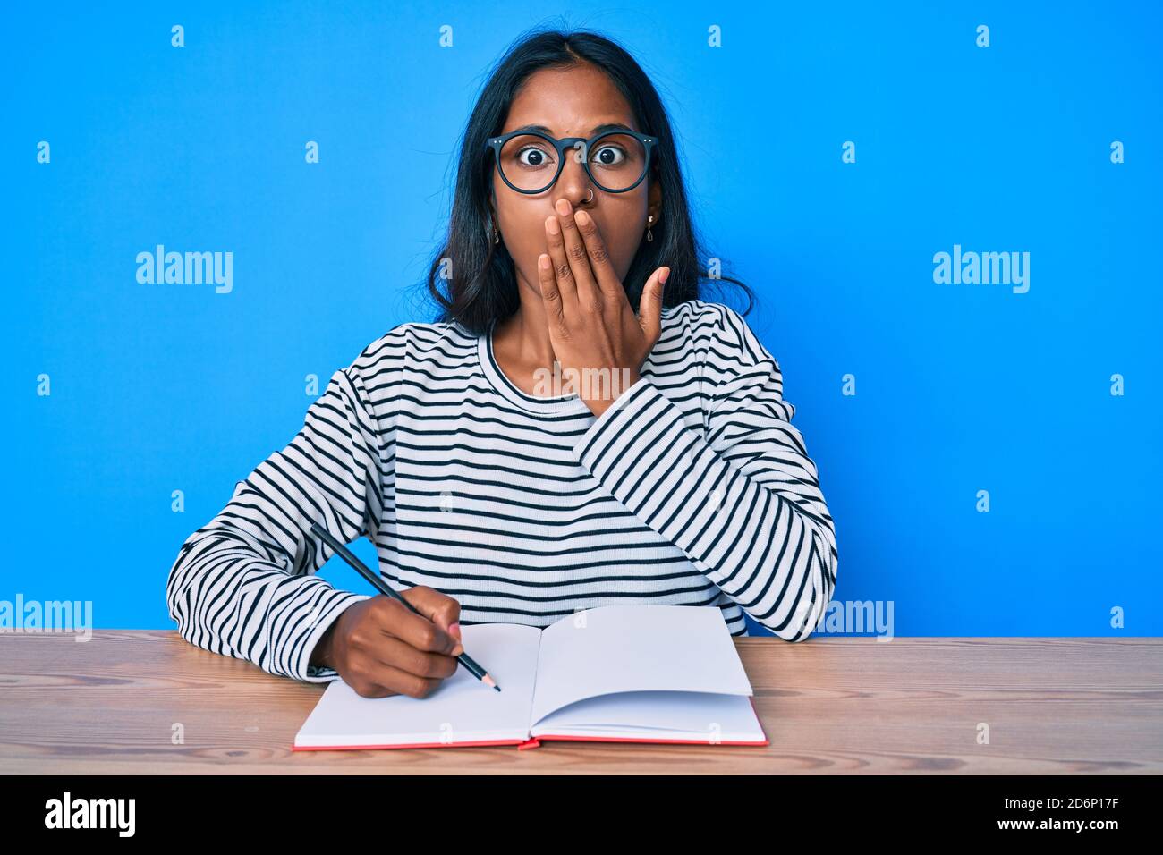 Young indian girl writing notebook sitting on the table covering mouth ...