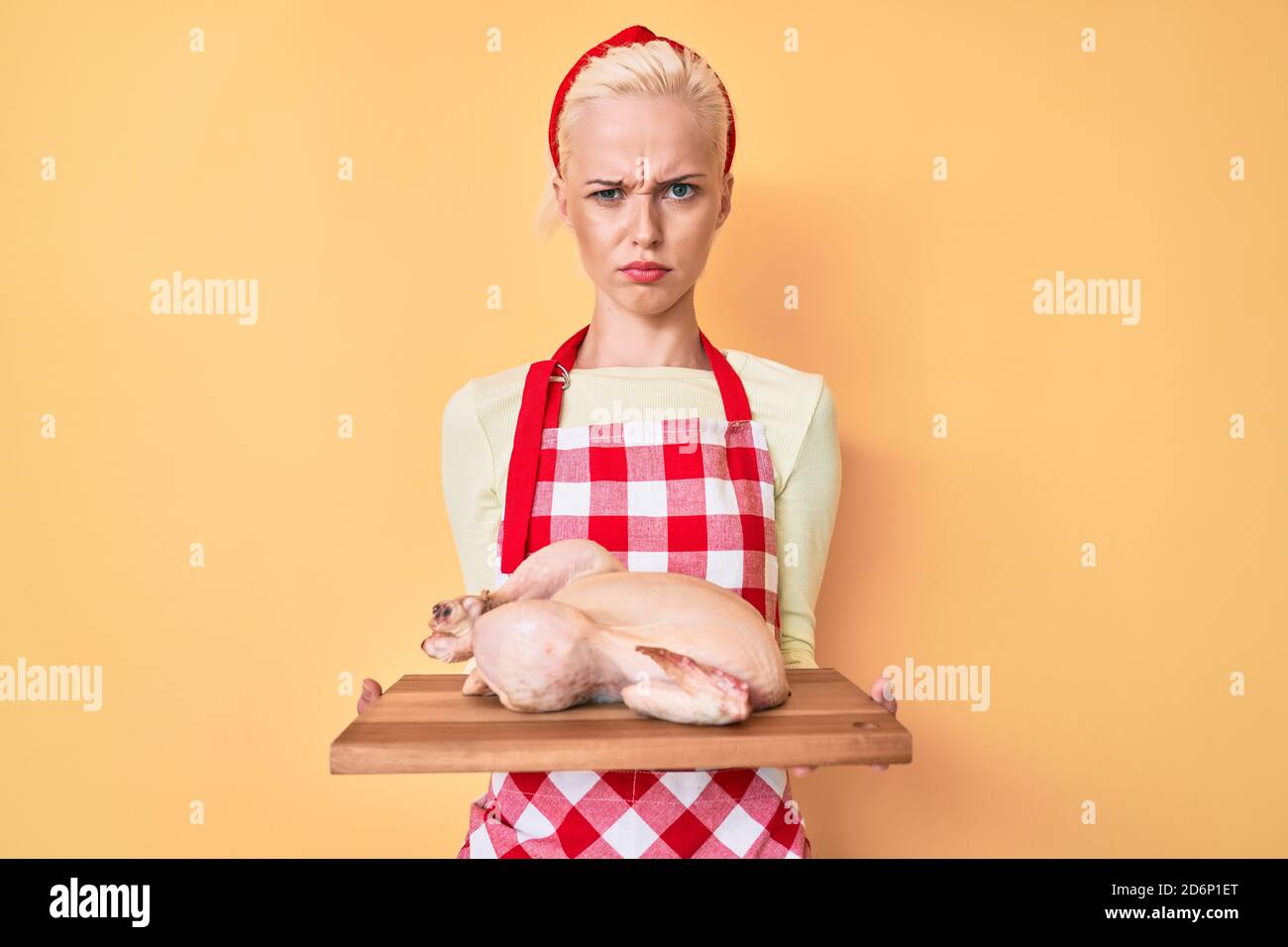 Young blonde woman wearing apron cooking chicken skeptic and nervous ...