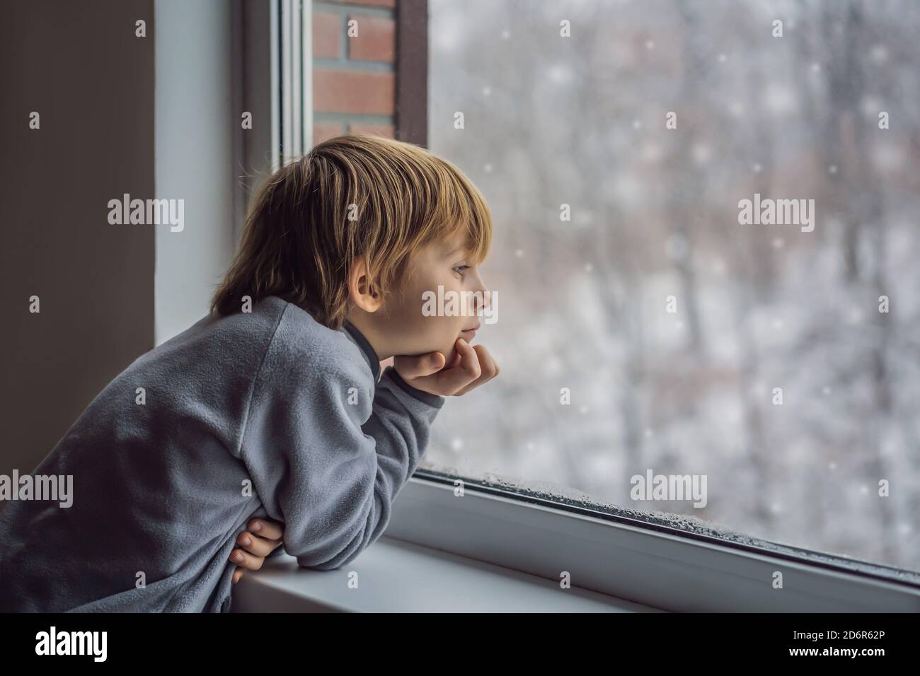 Happy adorable kid boy sitting near window and looking outside on snow ...