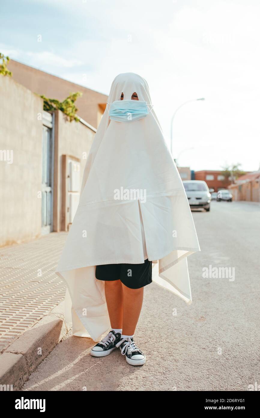 Unrecognizable kid in white ghost costume and with plastic jack o ...