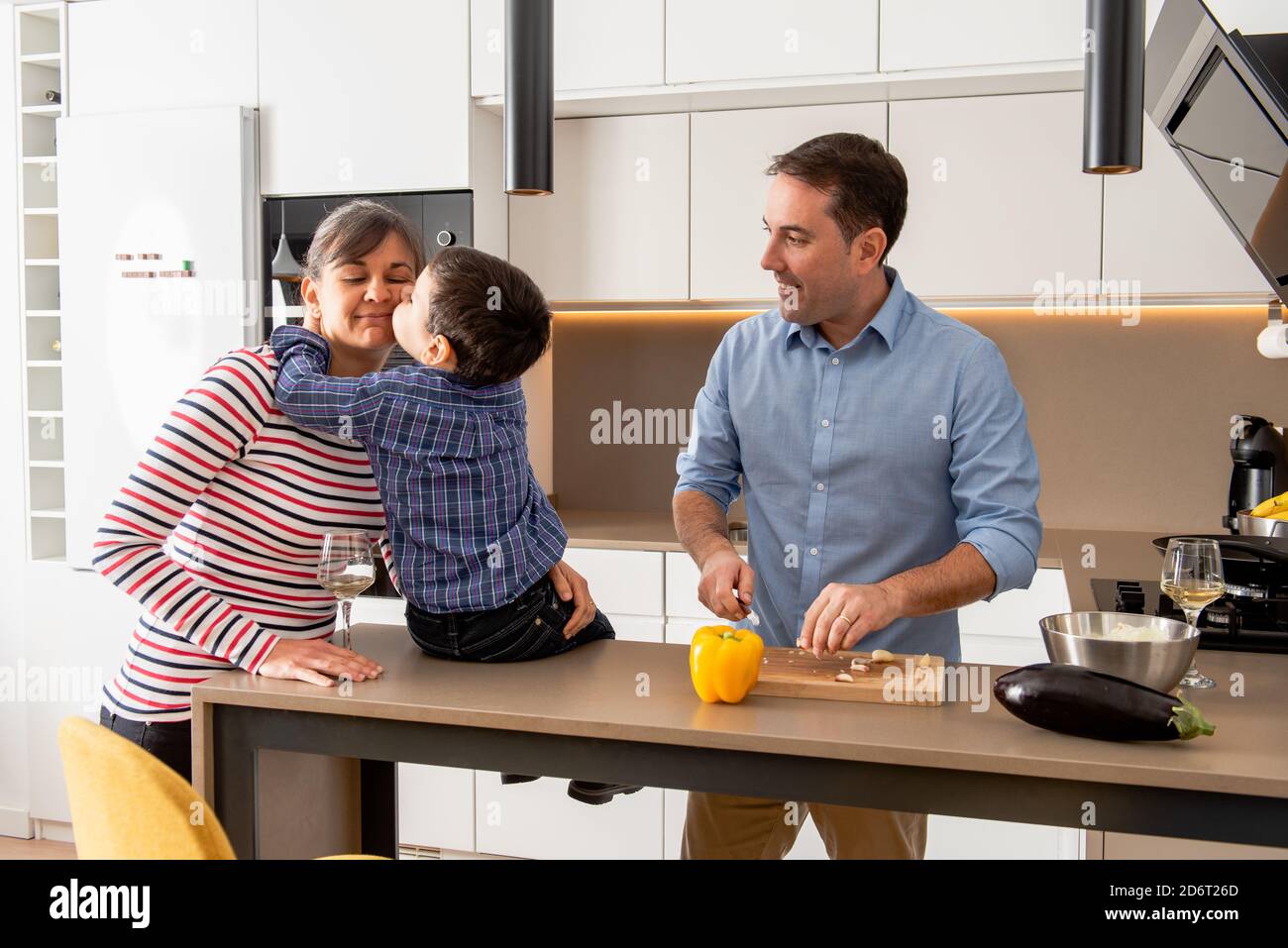 Cute little boy sitting on kitchen counter and kissing mothers cheeks ...