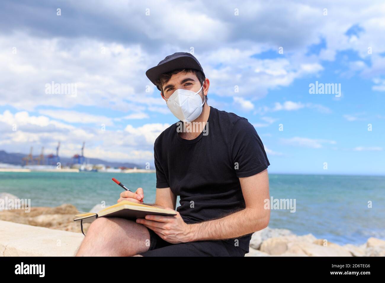 Male author in face mask and cap sitting with crossed legs on fence ...