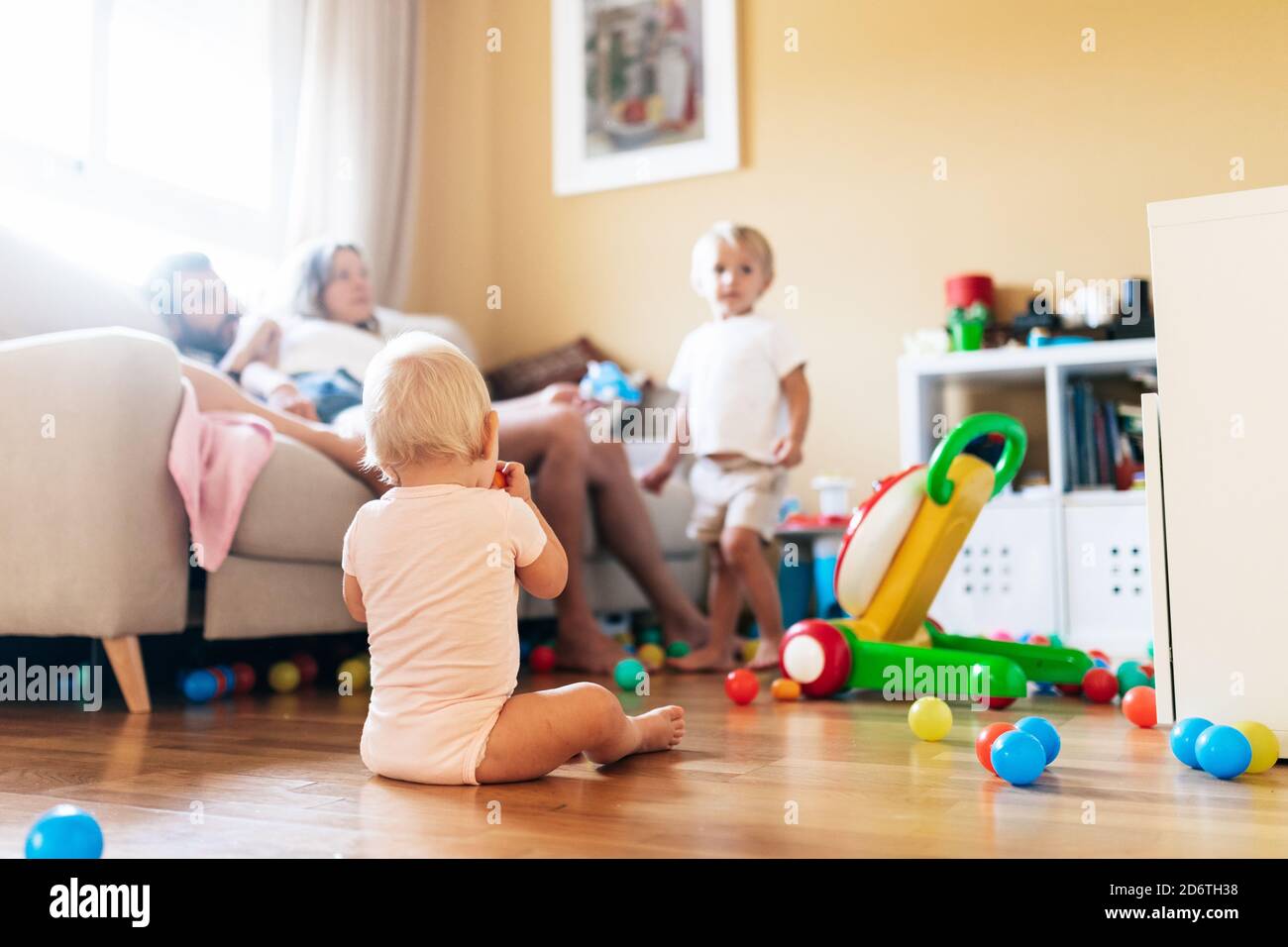 Back view of baby sitting on couch with toys spending time with brother ...