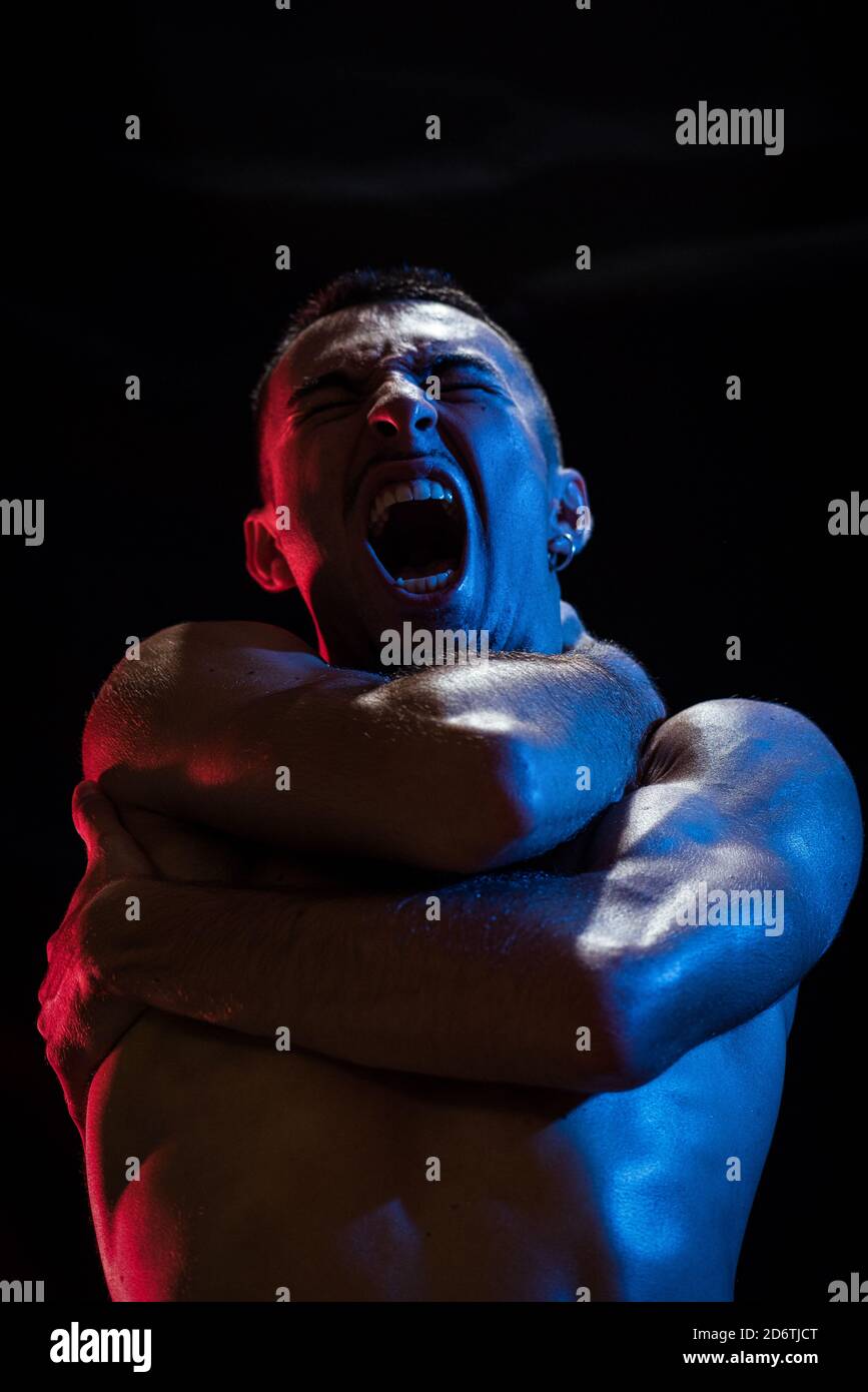 Unhappy shirtless sporty male shouting with eyes closed while embracing ...