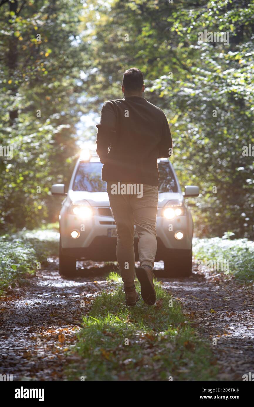 man running to car standing at the forest trail with headlights on ...
