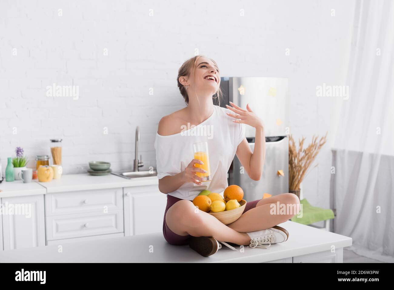 laughing woman in white t-shirt sitting on kitchen table with fruits ...