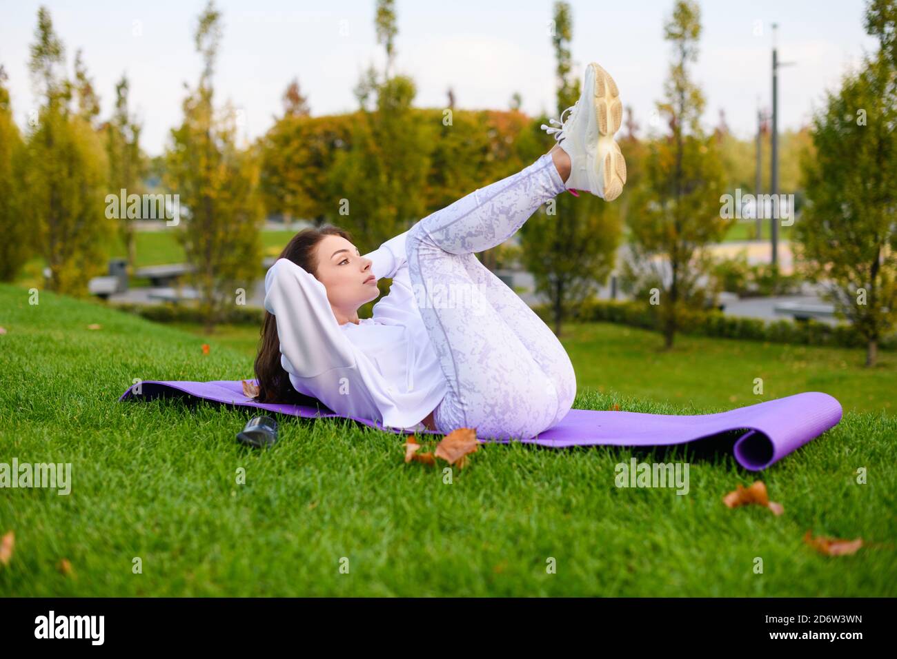 Sporty female in white clothes lay at mat outdoors and make stretching ...