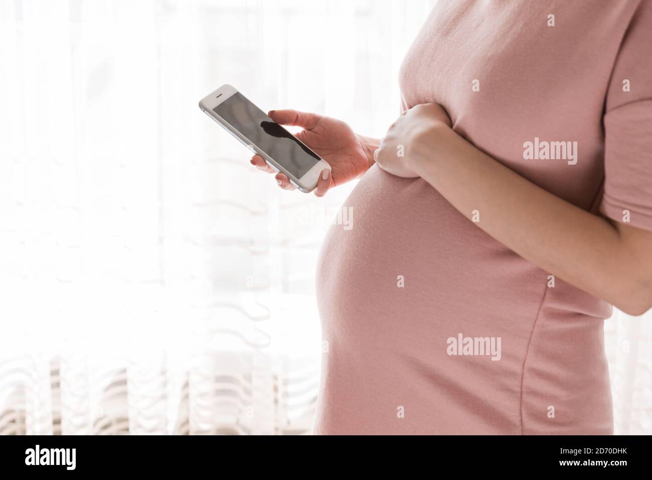 Pretty young pregnant woman standing by the window and texting message ...