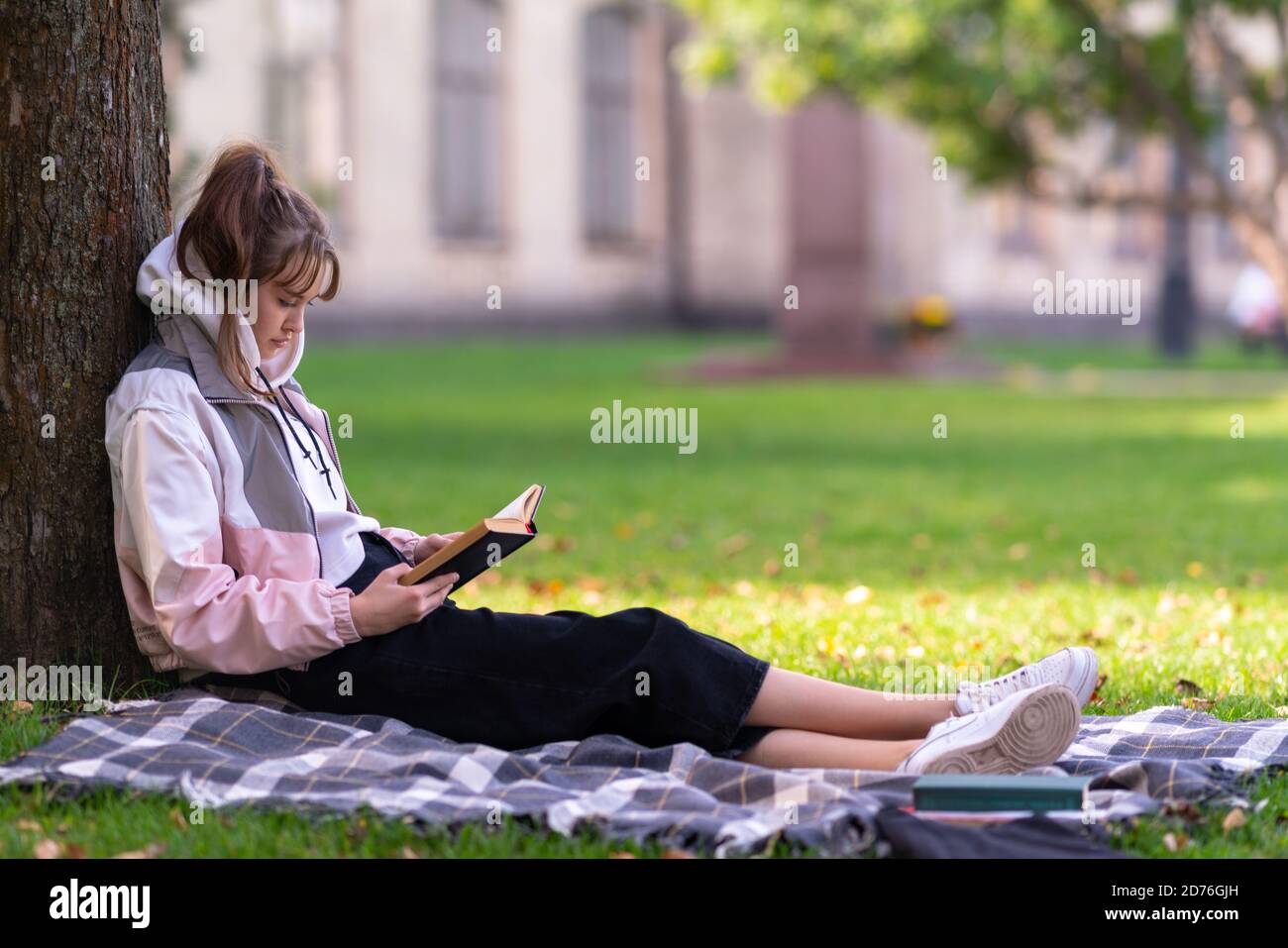 Young woman relaxing reading a book or studying under a shady tree in a ...