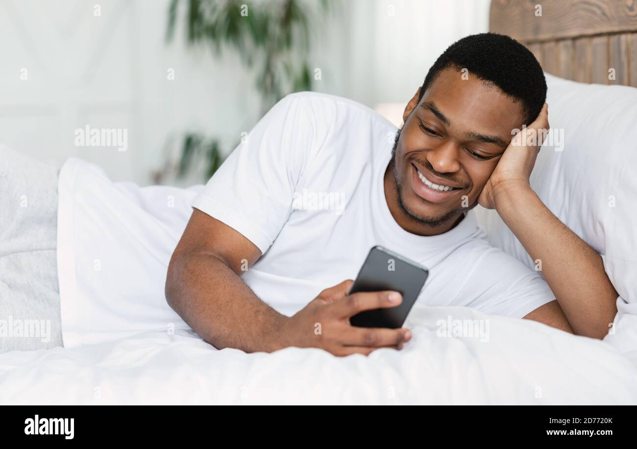 African Guy Using Cellphone Browsing Internet Lying In Bed Indoor Stock ...