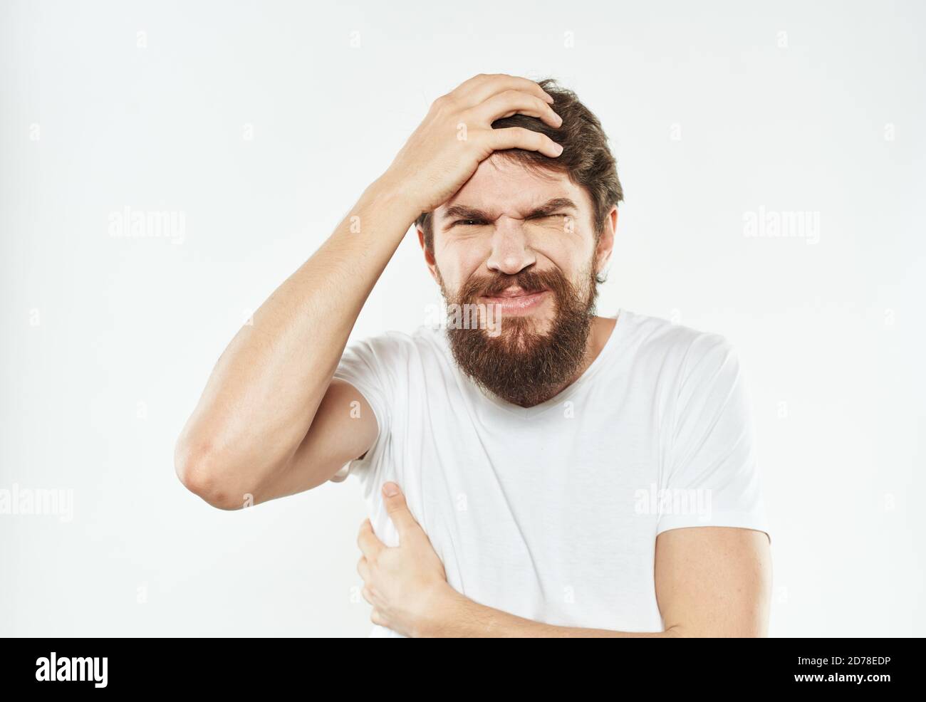 A puzzled man with a beard and in a white T-shirt on a light background ...
