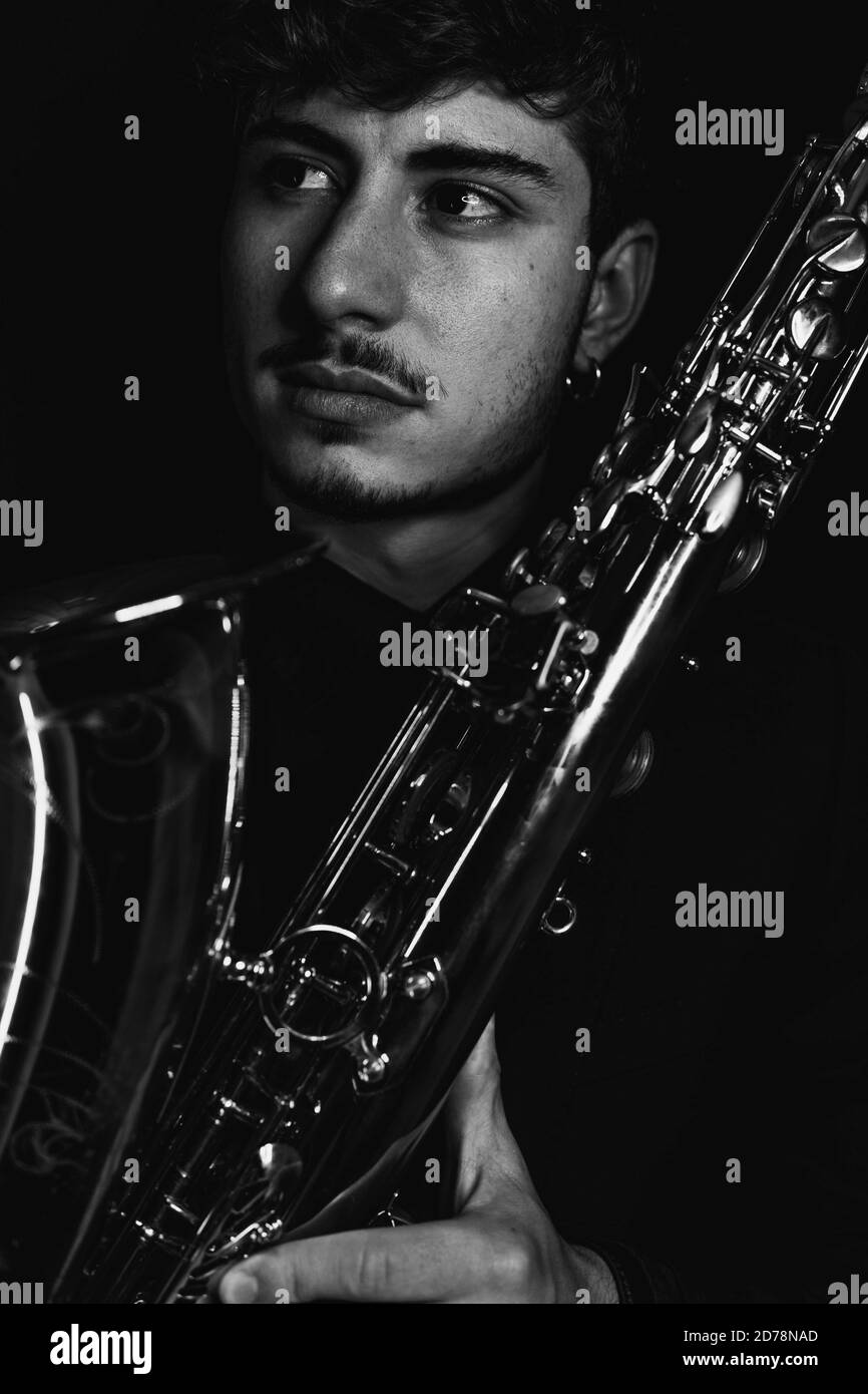 Grayscale shot of a handsome guy holding his saxophone on a dark ...