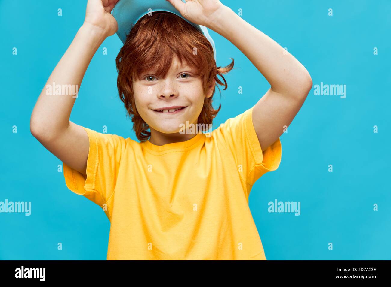 Cheerful red-haired boy bites his lower lip and holds on to his cap ...