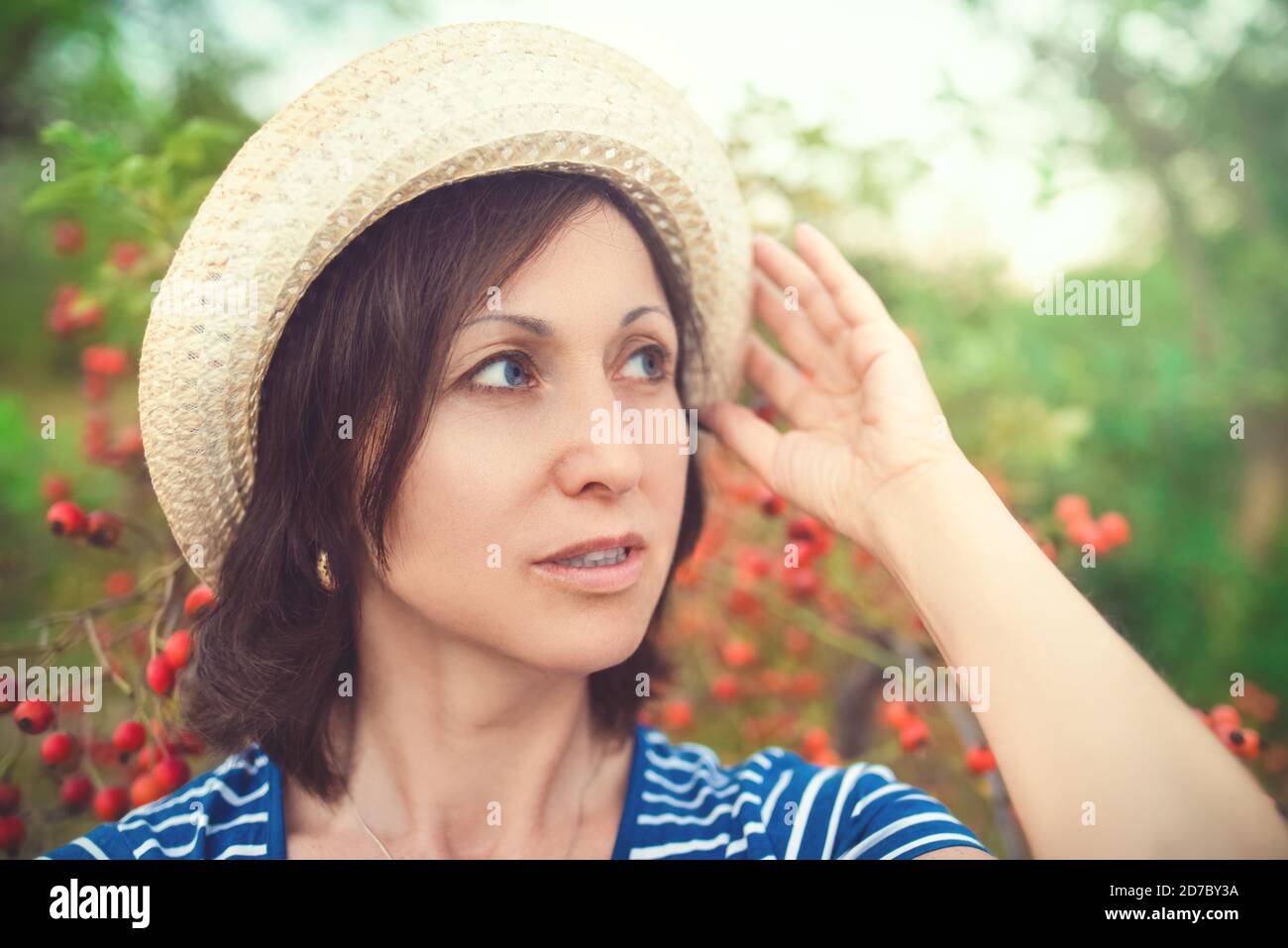 Image of beautiful middle-aged woman wearing straw hat laughing and looking at camera while ...