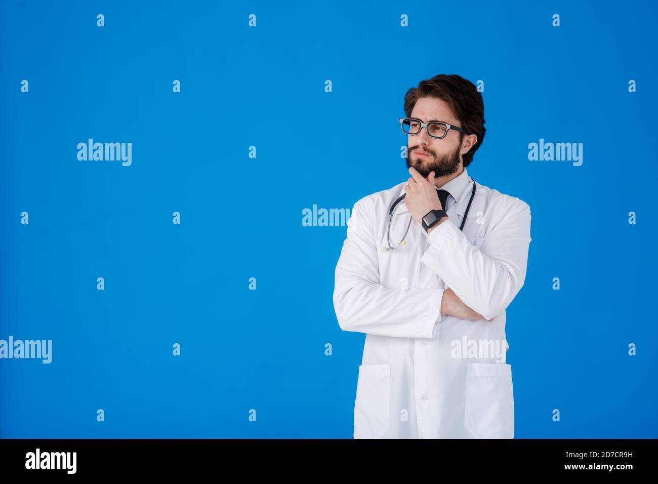 A young bearded doctor on a blue background in a white coat looks at ...