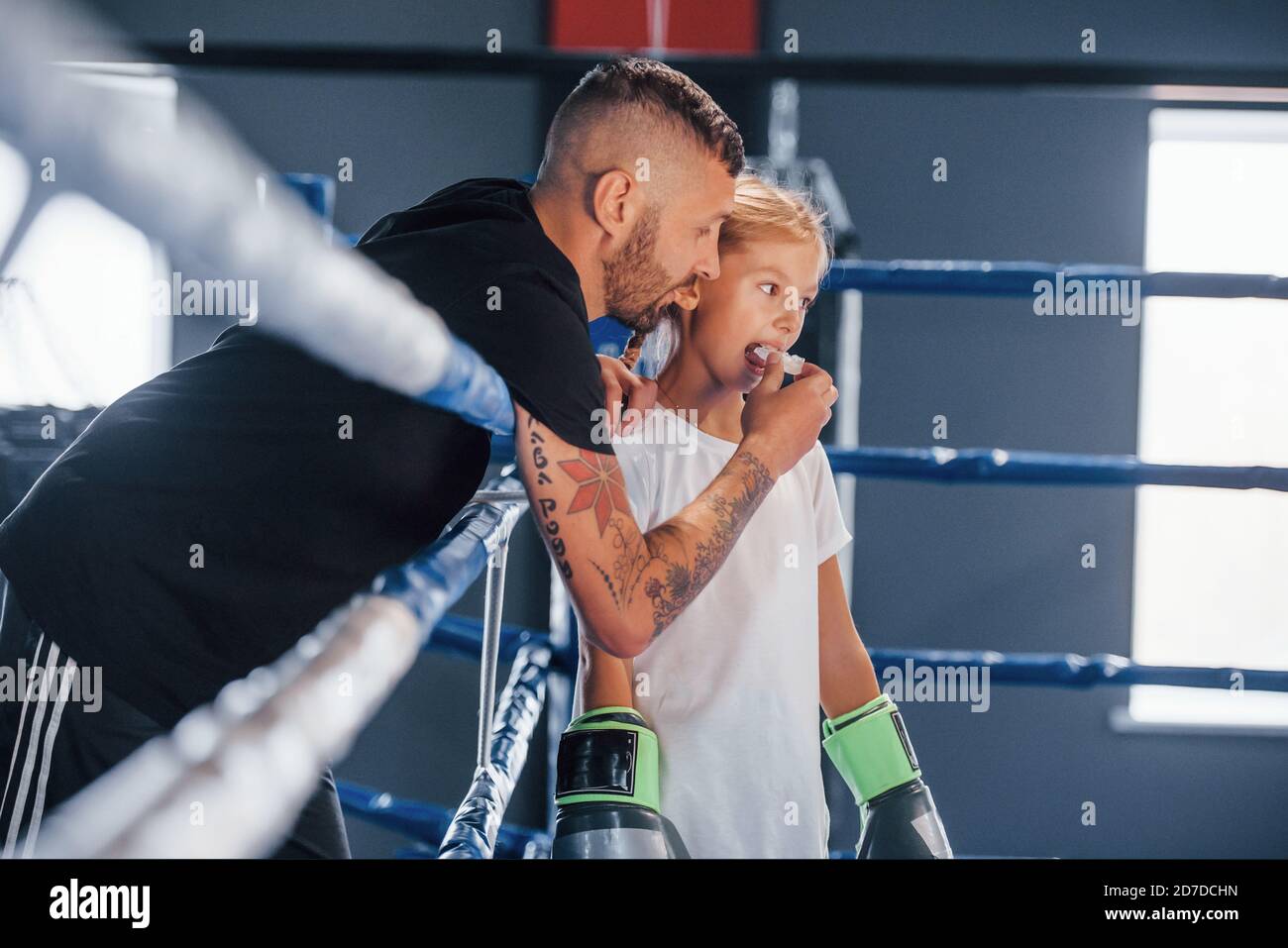Standing on the boxing ring. Young tattooed boxing coach teaches cute ...