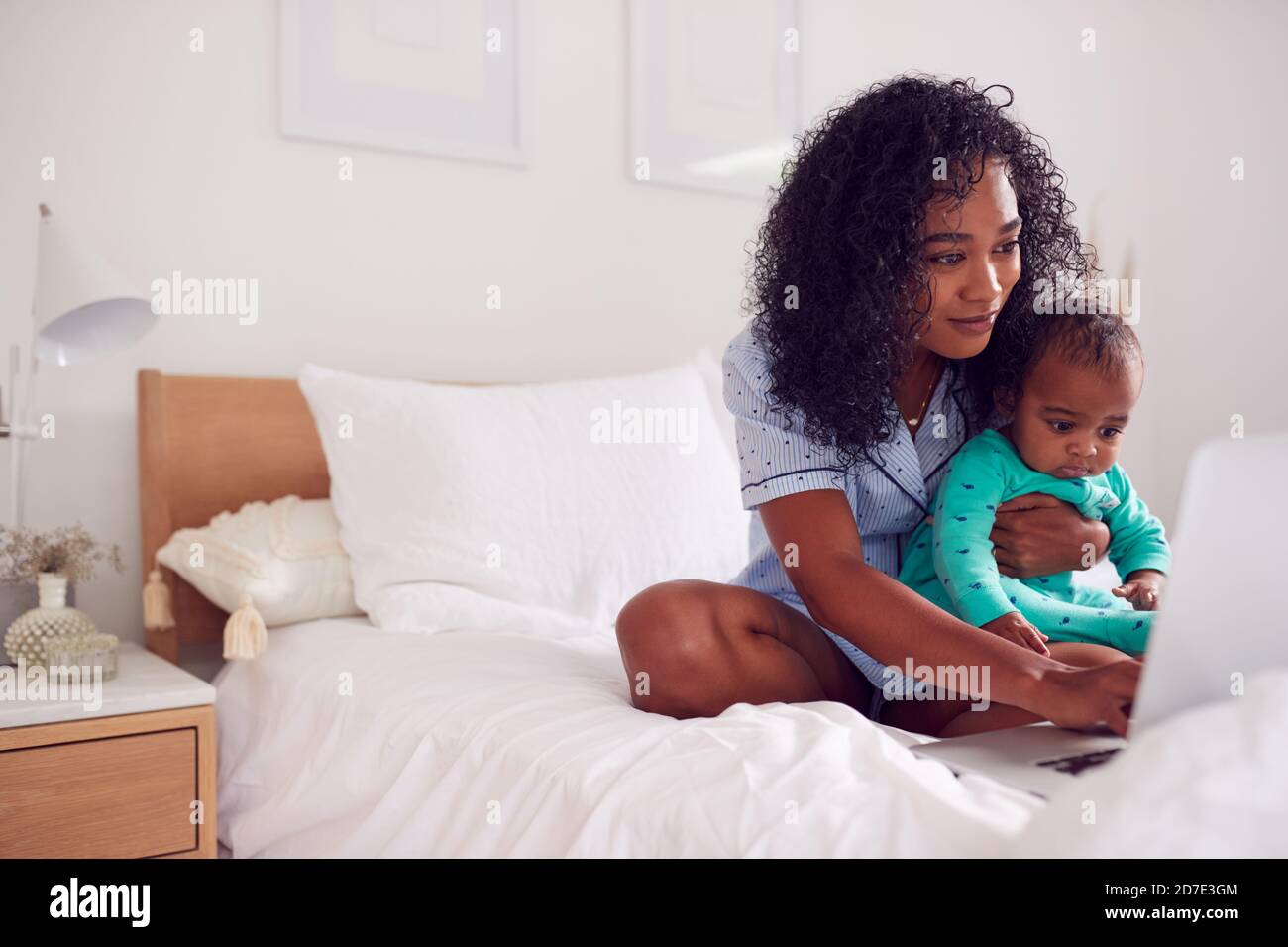 Mother Wearing Pyjamas With Baby Daughter In Bedroom Working From Home ...