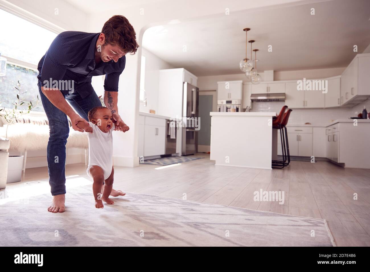 Parents Encouraging Smiling Baby Daughter To Take First Steps And Walk ...