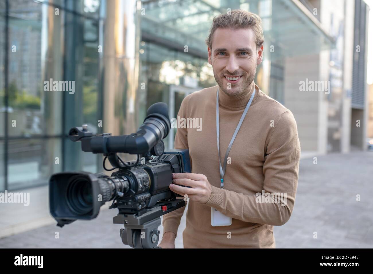 Fair-haired cameraman standing behind camera in front of modern ...