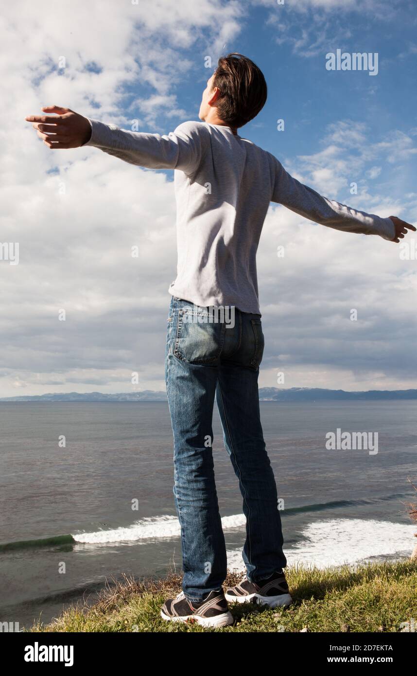 Blissful young man standing above the ocean view with both arms open ...