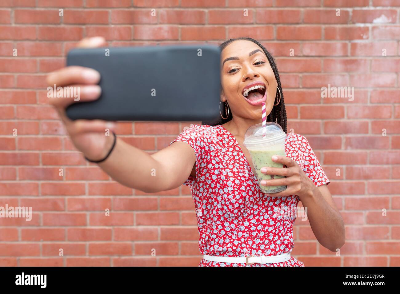Young Woman Taking Selfie While Drinking Smoothie Against Brick Wall