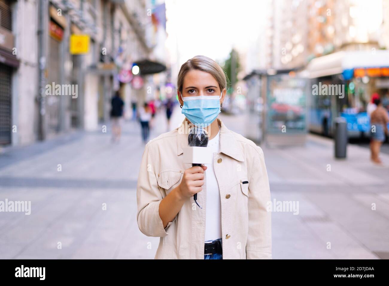 Female journalist wearing mask with microphone standing on street in ...