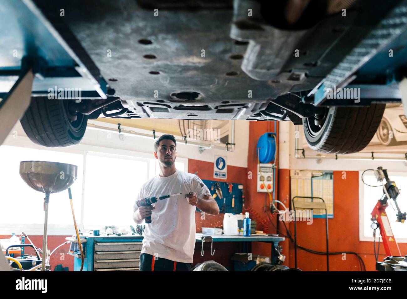 Young mechanic holding work tool looking at car while standing in auto ...