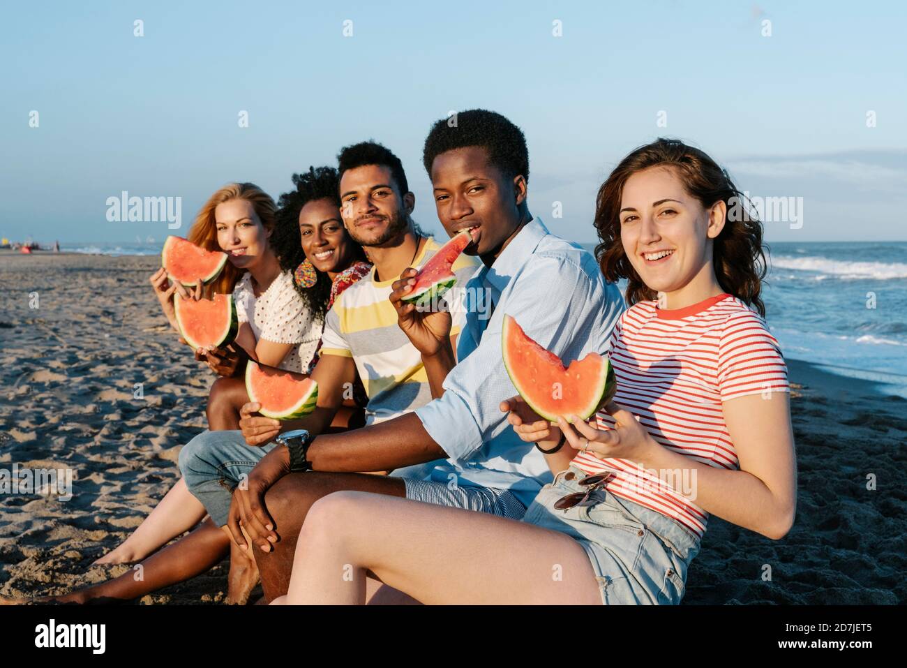 Smiling friends eating watermelon while sitting on beach during sunny ...