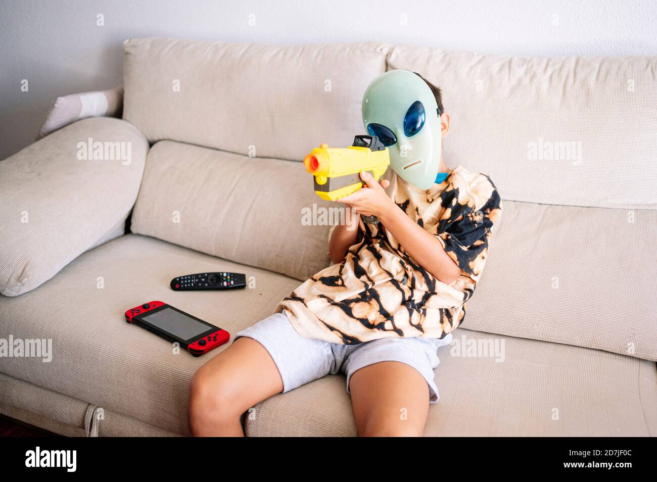 Boy wearing alien mask playing with toy gun while sitting on sofa at ...